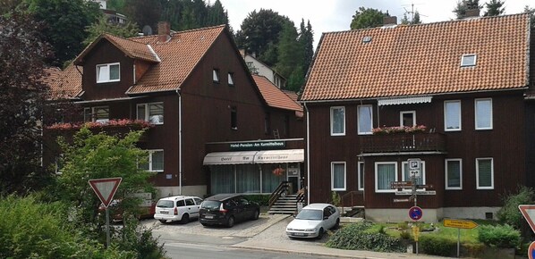 Um hotel tradicional europeu com telhado de telhas vermelhas, uma placa indicando que é um pensão, e um estacionamento com vários carros.