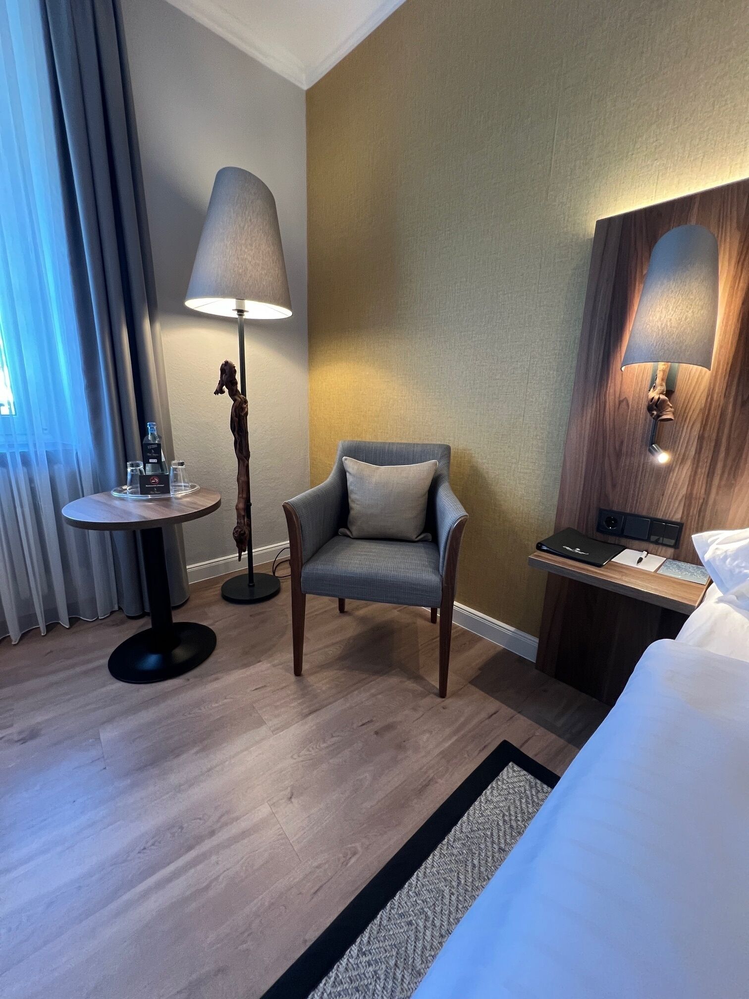 Quarto de hotel com uma cama, uma cadeira, uma mesa redonda com abajur e uma mesinha de cabeceira também com abajur.