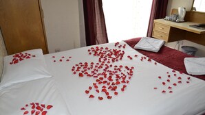 Quarto de hotel com cama coberta de pétalas de rosa, escrivaninha de madeira com chaleira e janela com cortinas.