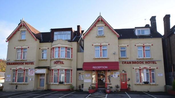 Um edifício de três andares com o nome "Cranbrook Hotel" na fachada. Possui um toldo vermelho sobre a entrada, onde se lê "Recepção". O edifício tem várias janelas com detalhes vermelhos e um telhado plano.