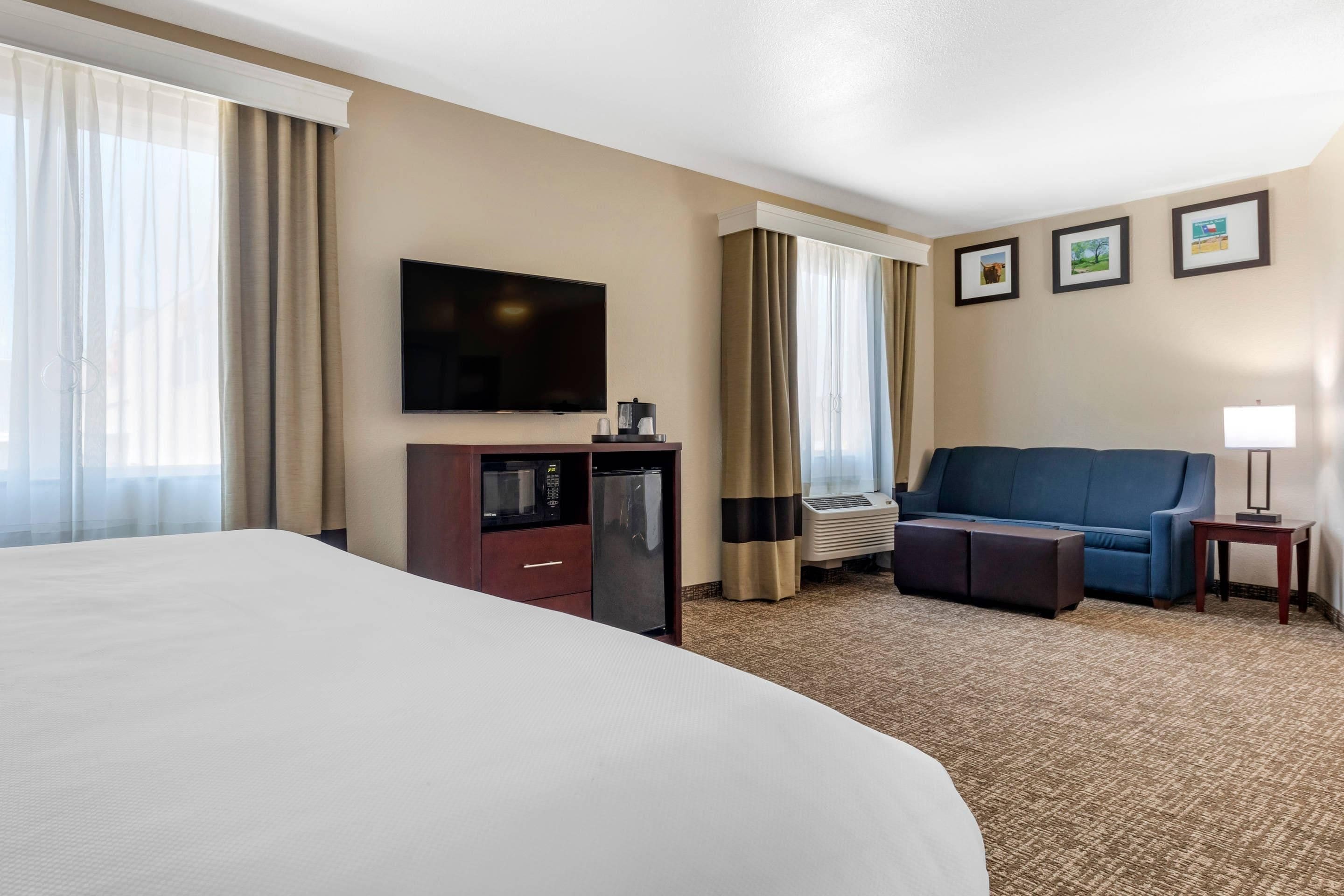 Quarto de hotel com uma cama grande, TV de tela plana, minibar, sofá e uma pequena mesa com luminária.