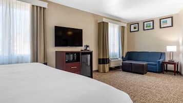 Quarto de hotel com uma cama grande, TV de tela plana, minibar, sofá e uma pequena mesa com luminária.