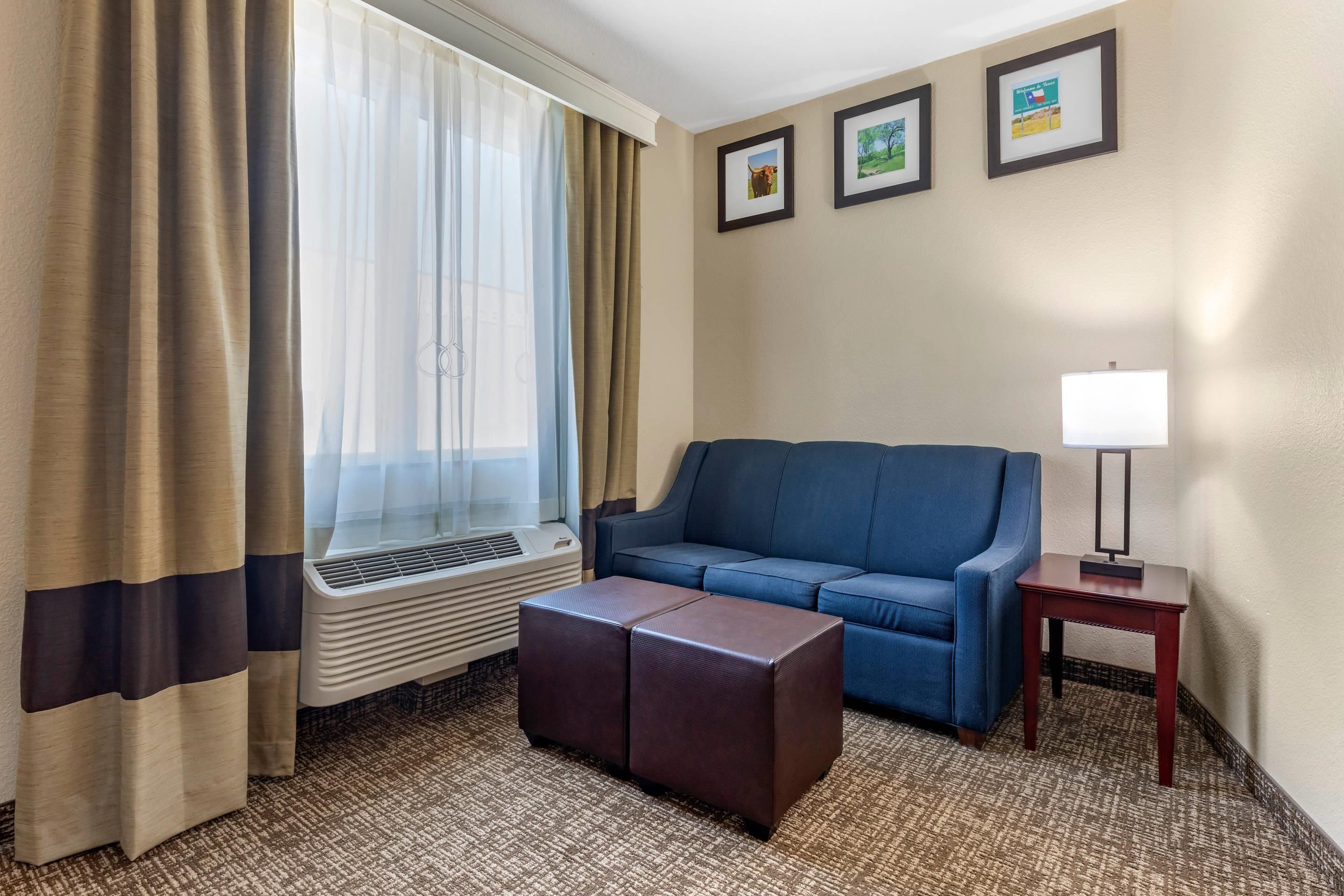 Quarto de hotel com sofá azul, um banquinho marrom, uma mesa pequena com abajur, janela com cortinas e três quadros na parede.