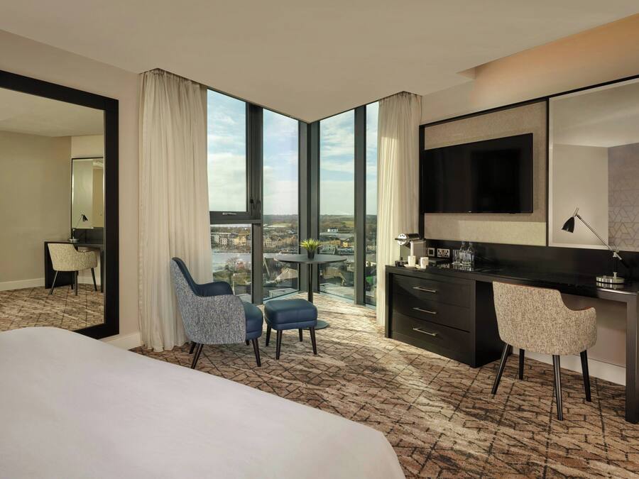 Quarto de hotel com uma cama grande, uma mesa com cadeira, televisão e vista para a cidade através de amplas janelas.