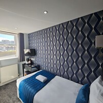Quarto de hotel com uma cama grande, uma escrivaninha com abajur, uma janela com vista para edifícios e parede com padrão.