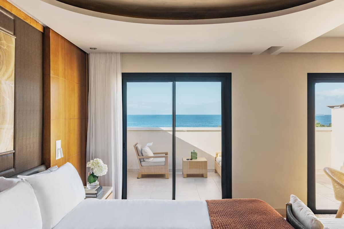 Quarto de hotel com uma cama, uma pequena mesa com um vaso de flores, uma cadeira e vista para a praia através de uma porta deslizante.