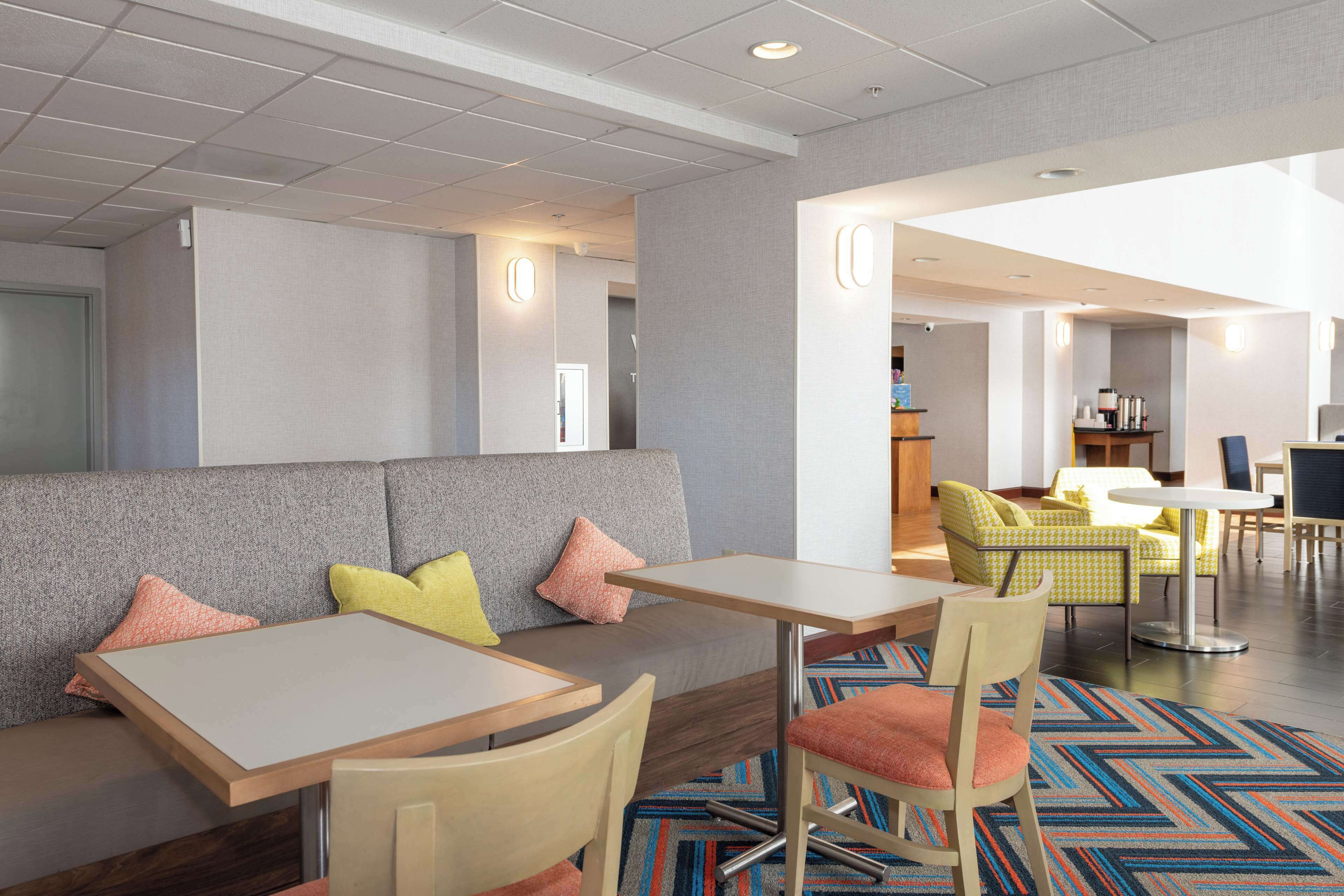 Um hall de hotel moderno com um sofá cinza, mesas e cadeiras de madeira, e um tapete colorido.