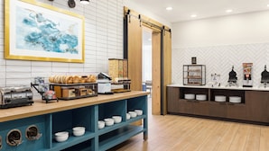 Uma mesa de pequeno-almoço com máquina de café, torradeira e pão exposto, um quadro na parede e um revestimento de parede em padrão herringbone.