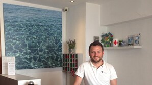 Uma receção com acabamento em madeira, uma janela ampla com vista para águas límpidas e uma estante com vários objetos, incluindo uma planta e um kit de primeiros socorros.