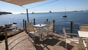 Área externa com mesas e cadeiras sob um toldo, com vista para um mar calmo, barcos e uma linha de costa distante.