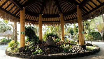 Um pavilhão coberto com telhado de palha, rodeado por um jardim com pedras e plantas tropicais.