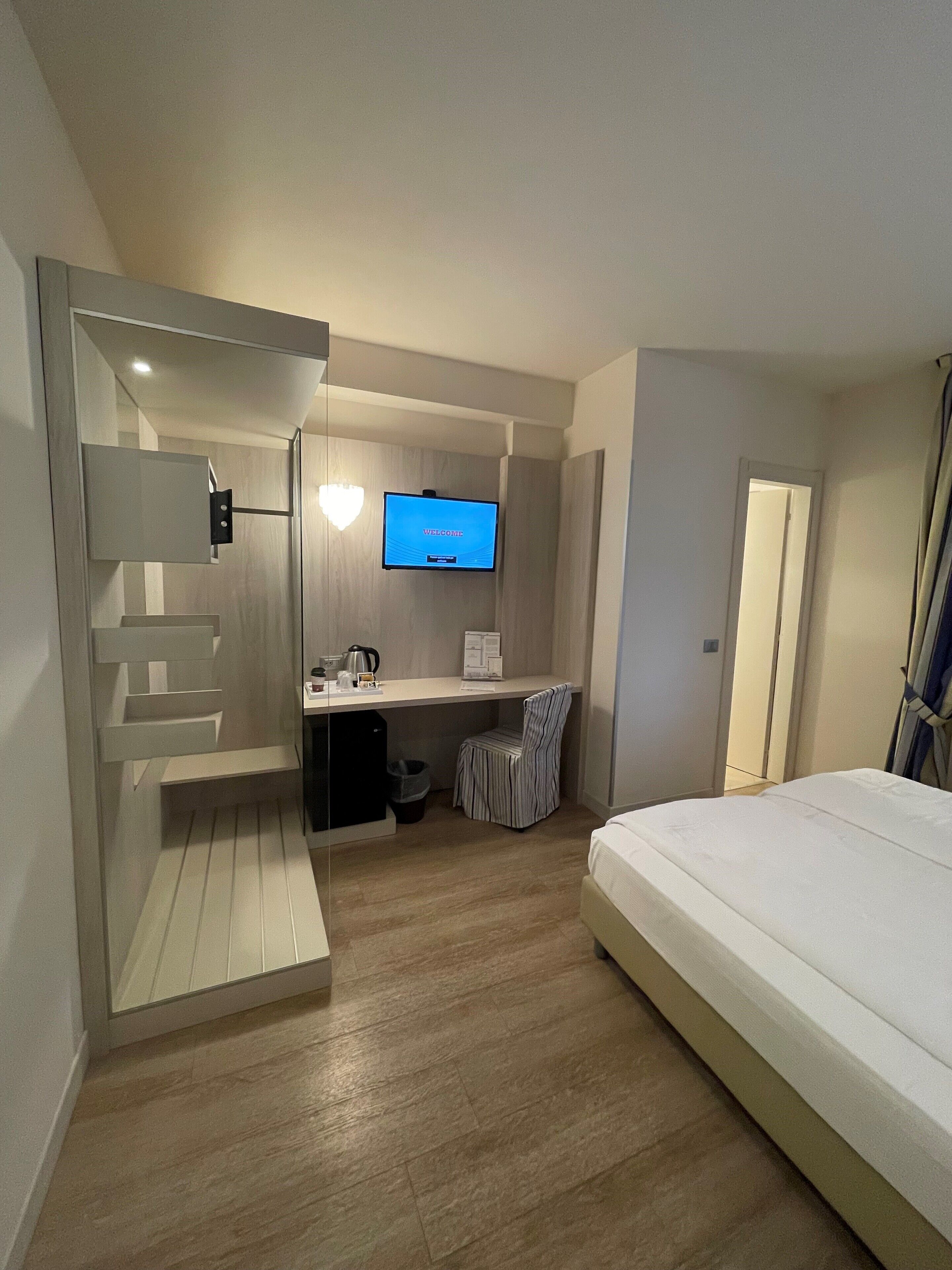 Quarto de hotel compacto com cama, escrivaninha, cadeira, televisão e pequeno espaço de cozinha.