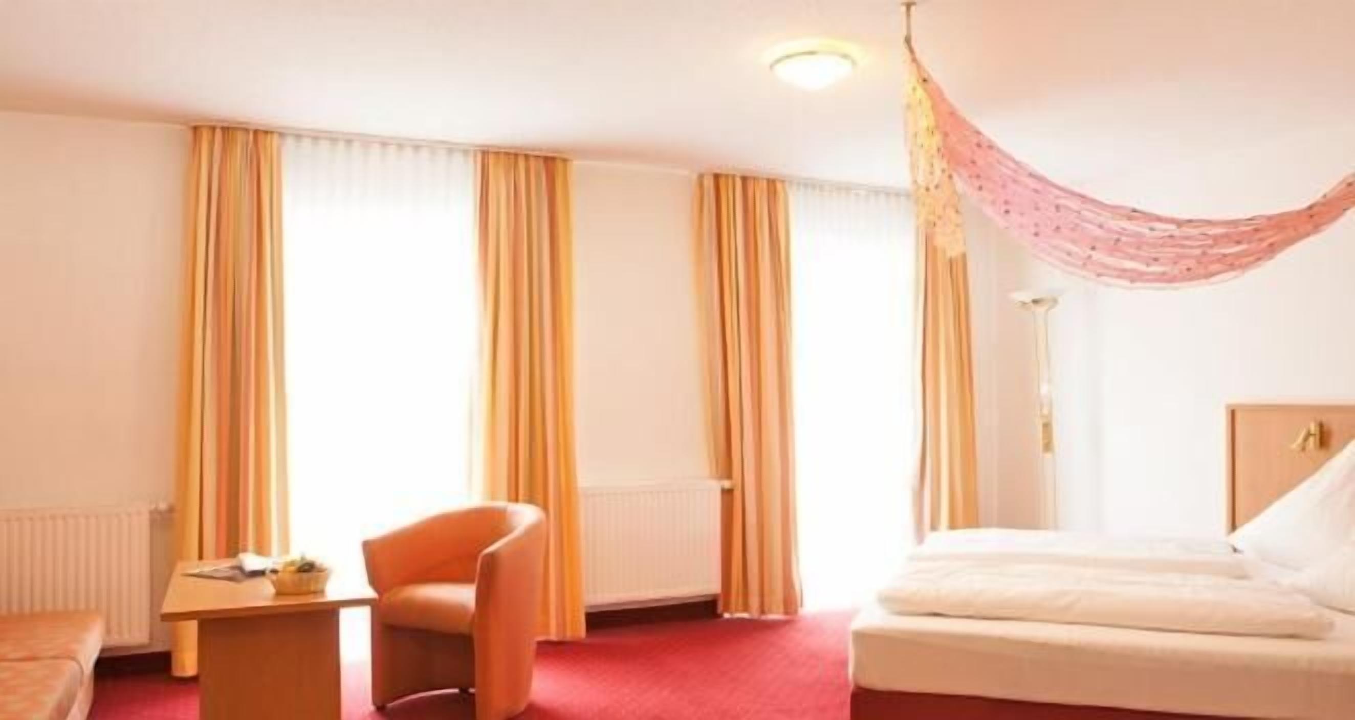 Quarto de hotel com cama, escrivaninha, cadeira e uma janela grande com cortinas.