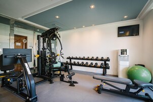 Uma academia com equipamentos de ginástica, um banco, uma bola verde para exercícios e uma televisão na parede.
