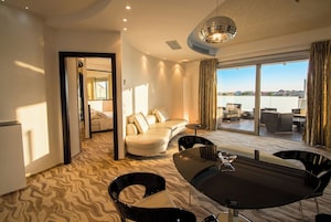 A hotel room with a sofa, a dining table with chairs, a large window with curtains, and a ceiling light fixture.
