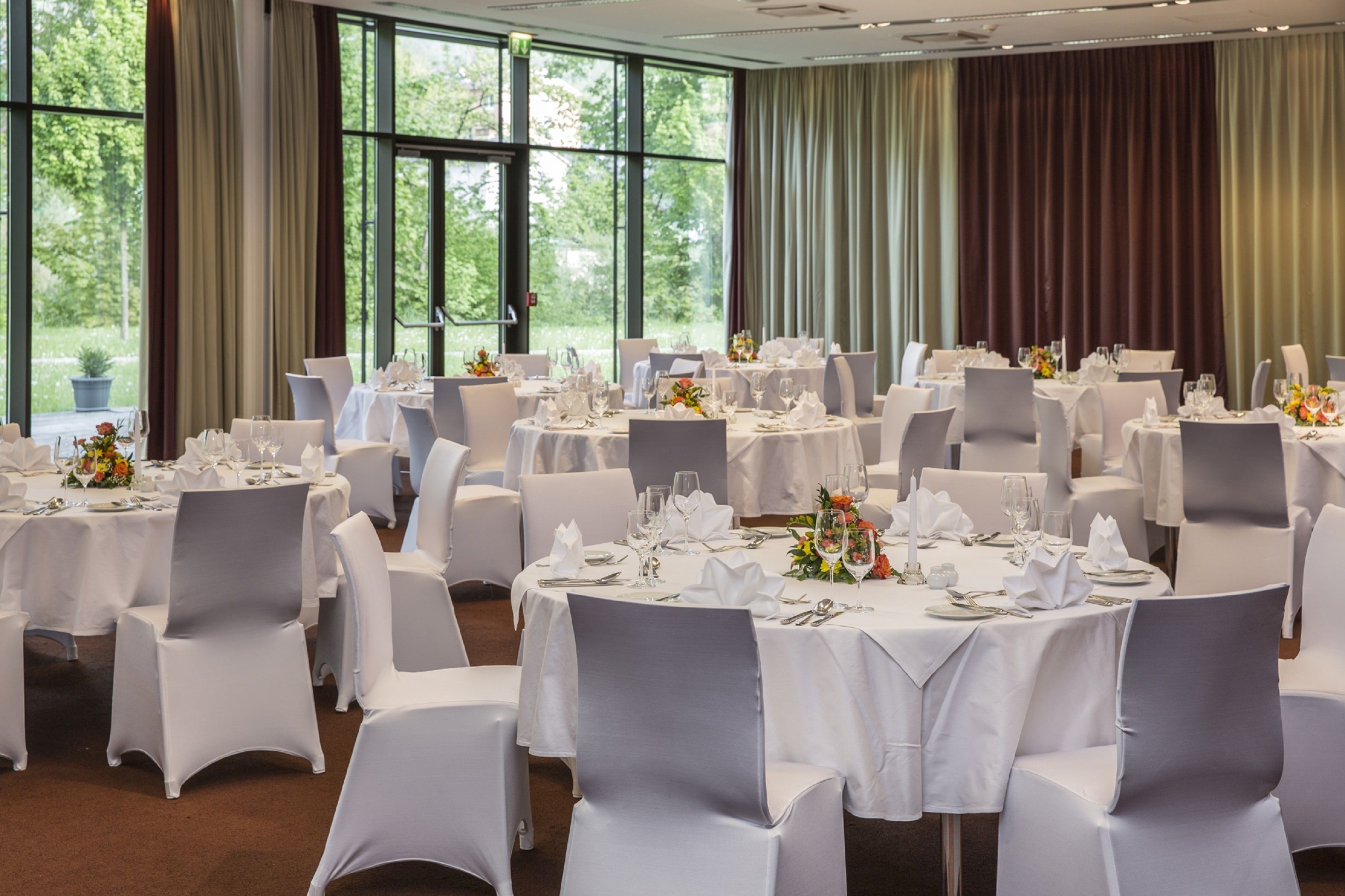 Um salão de banquetes com mesas redondas dispostas para refeições, grandes janelas com cortinas e vista para o exterior arborizado.