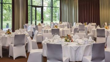 Um salão de banquetes com mesas redondas dispostas para refeições, grandes janelas com cortinas e vista para o exterior arborizado.
