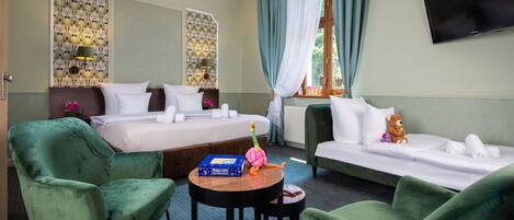 Quarto de hotel com duas camas, um sofá, poltronas verdes, uma mesa redonda pequena e uma televisão na parede.