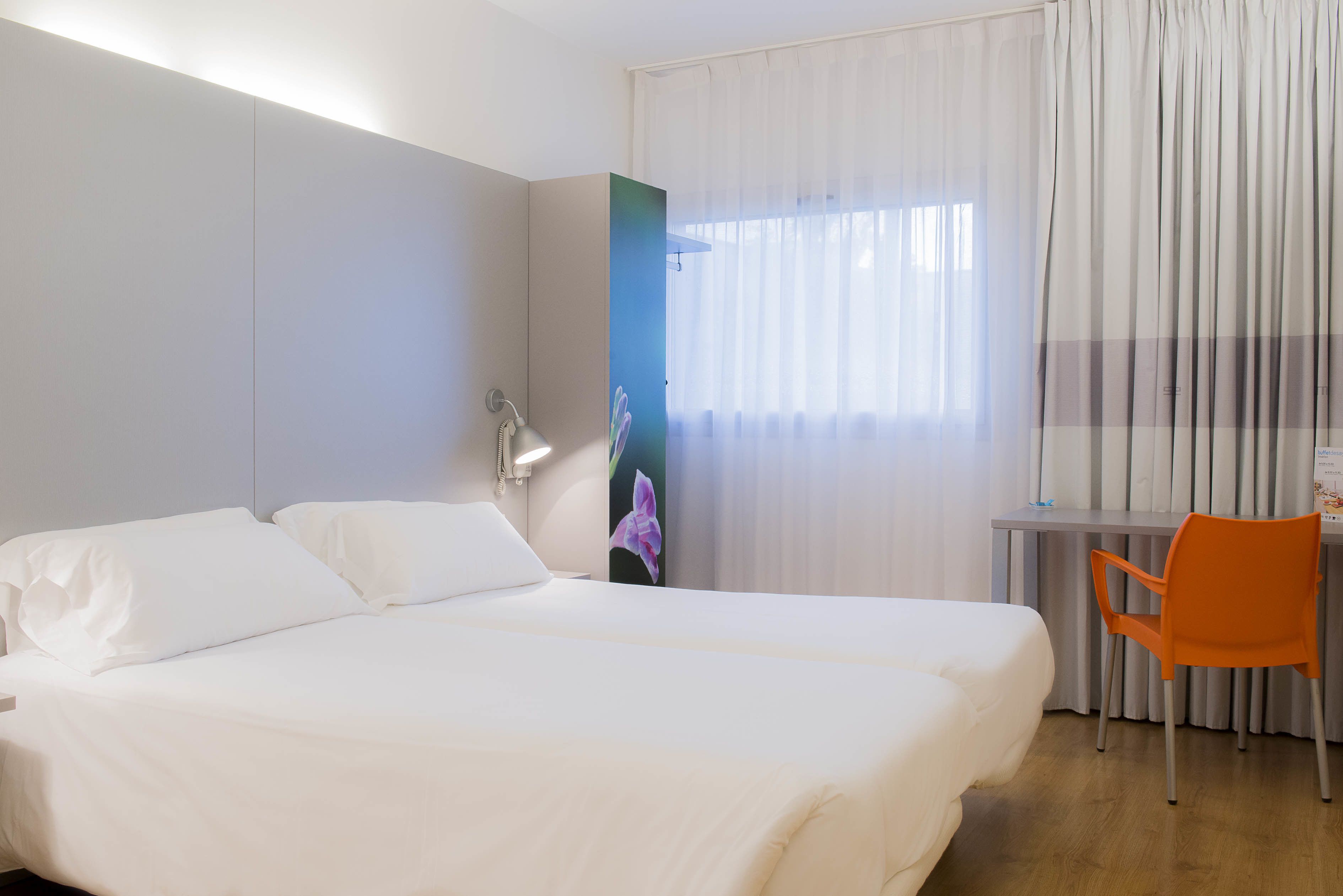 Quarto de hotel com duas camas, uma escrivaninha, uma cadeira laranja e uma janela com cortinas translúcidas.