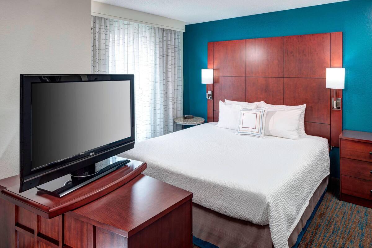 Quarto de hotel com uma grande TV de tela plana sobre um suporte de madeira, uma cama com roupa de cama branca, uma parede azul de destaque e duas luminárias de cabeceira.