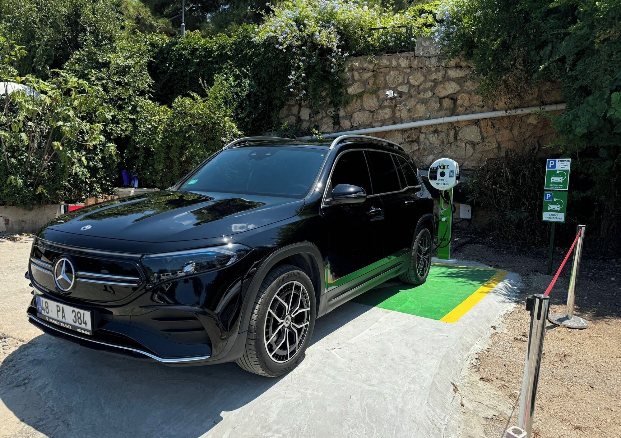 Um SUV preto estacionado numa estação de carregamento para veículos elétricos, com um muro de pedra e vegetação ao fundo.
