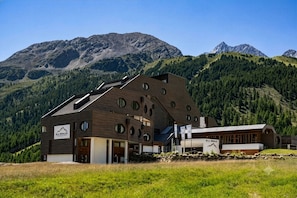 Um edifício moderno com telhado inclinado e janelas circulares, cercado por um campo gramado e montanhas.