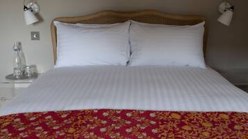 Cama bem arrumada com lençóis brancos e colcha floral vermelha. Há duas luminárias de cabeceira, uma de cada lado da cama.