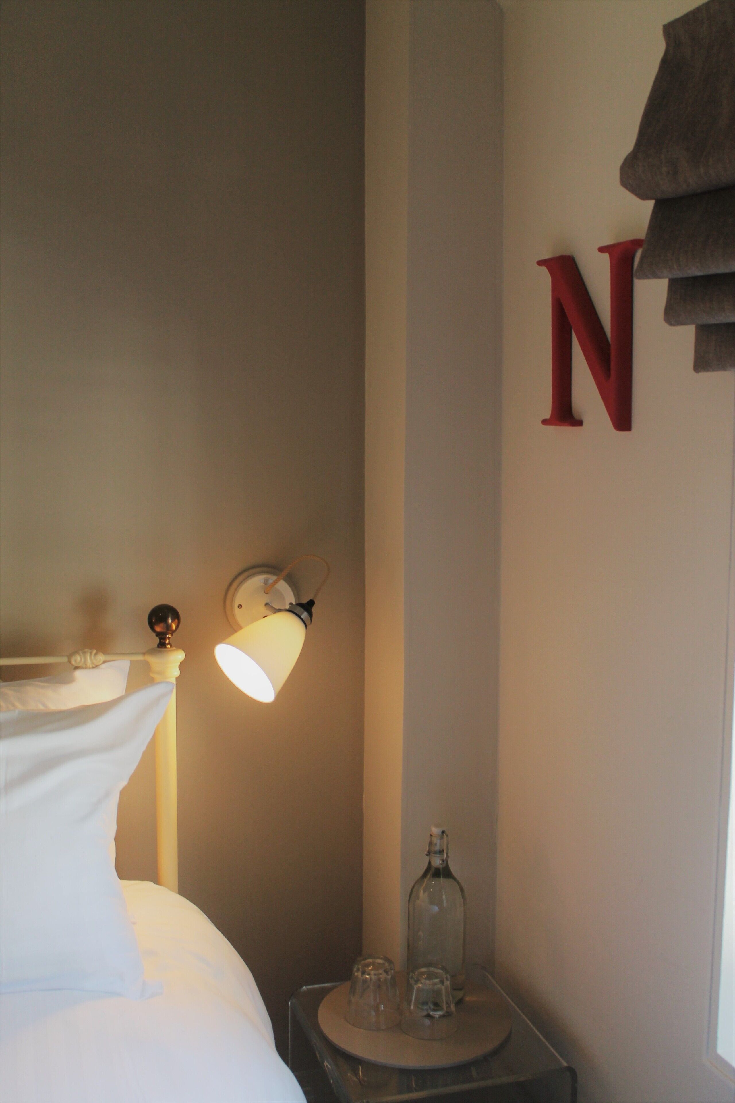 Cama bem arrumada com uma luminária, uma garrafa e copos em uma mesa de cabeceira, e a letra 'N' na parede.