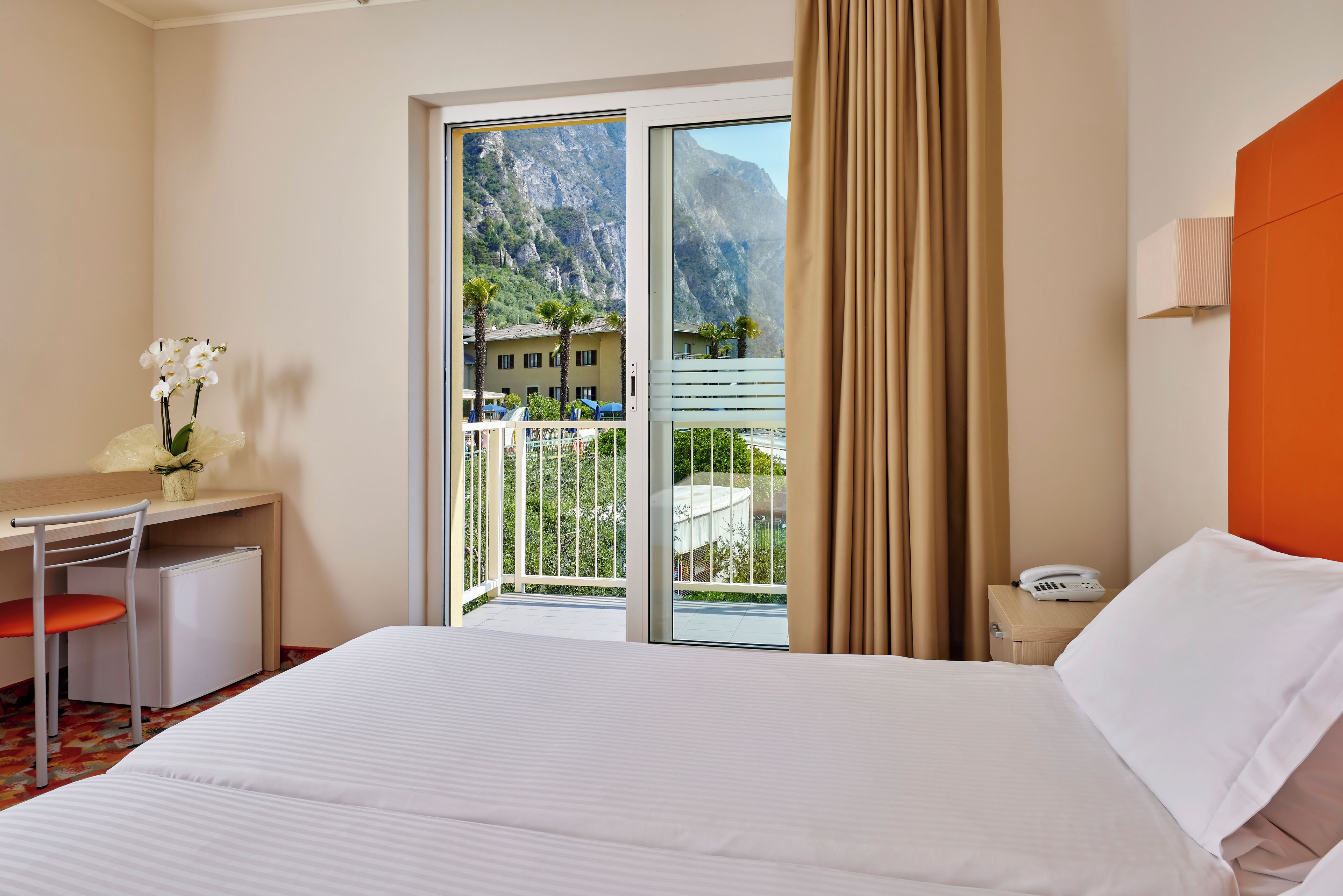 Quarto de hotel com cama, escrivaninha com cadeira, pequena geladeira, telefone e vista para uma montanha e um edifício através de uma porta de vidro.