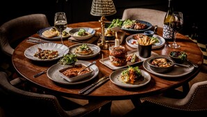 Mesa de jantar redonda posta com diversos pratos, travessas e talheres, acompanhada de uma luminária e taças de vinho.