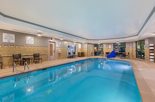 Piscina interna com aviso de proibição de saltos, placa indicando presença de salva-vidas e área ao redor da piscina com cadeiras e um espaço coberto para sentar.