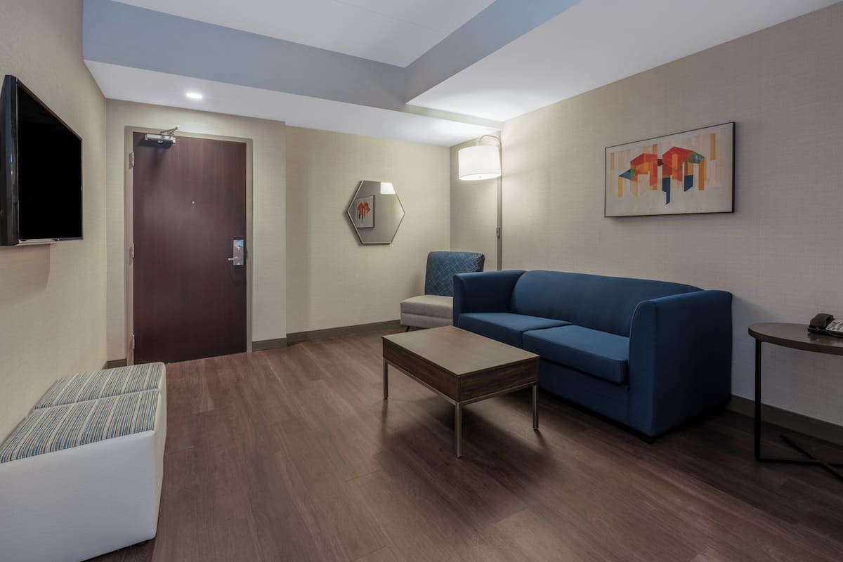 Quarto de hotel moderno com piso de madeira, sofá azul, mesa pequena, TV fixada na parede e um quadro.