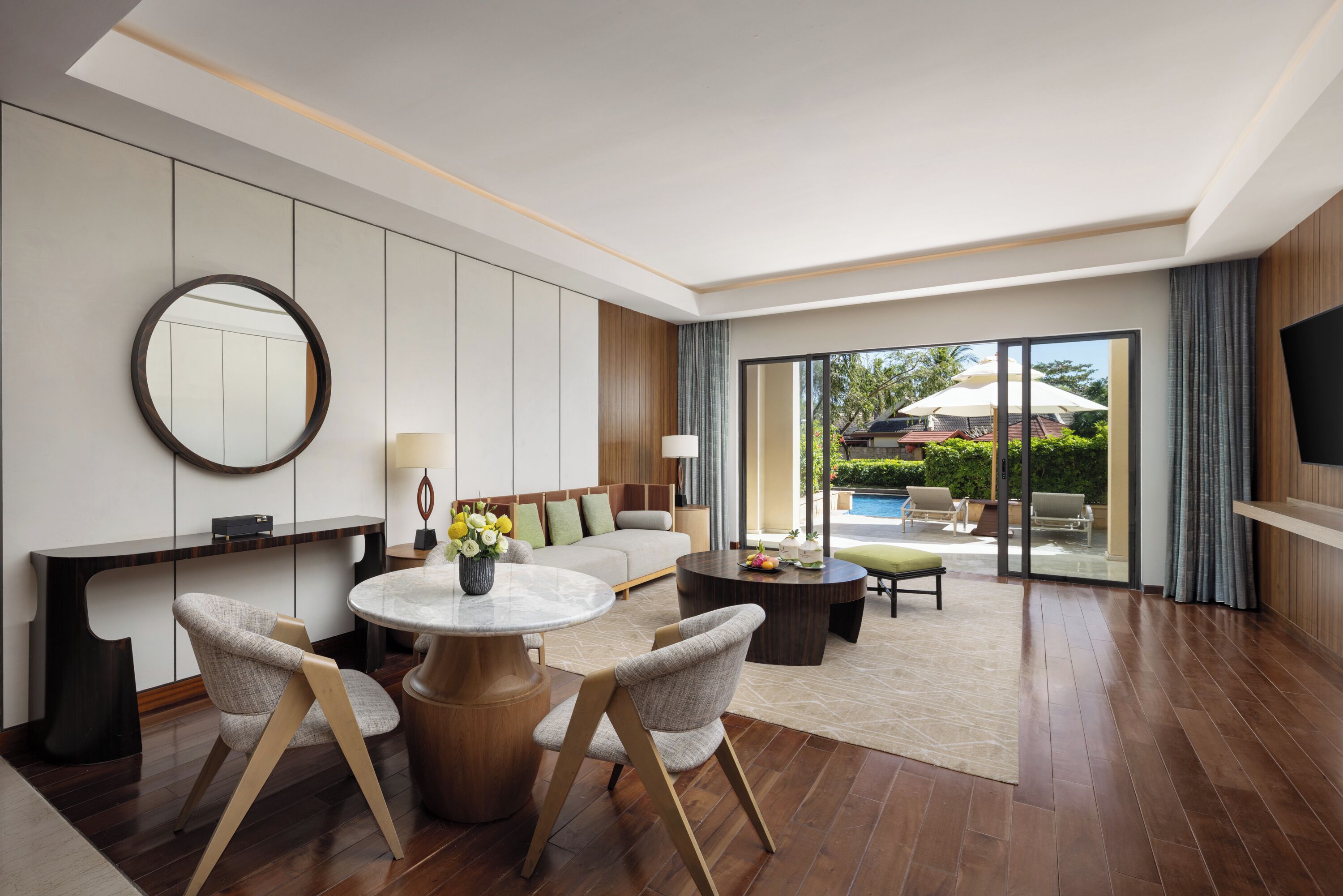 Quarto de hotel moderno com área de refeições, um sofá, televisão e vista para a área da piscina externa.
