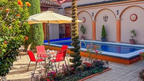 Uma área à beira da piscina com um terraço ladrilhado, cadeiras vermelhas, um guarda-sol amarelo e plantas em vasos.