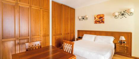 Quarto de hotel com cama e mesa de madeira, e uma cadeira. Há uma mesinha de cabeceira com abajur e um ponto de luz na parede.