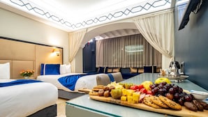 Quarto de hotel com duas camas, uma mesa de jantar com tampo de vidro e uma tábua de frutas e queijos, e vista da cidade através de grandes janelas.