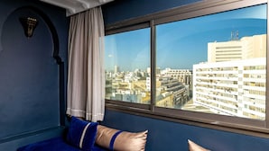 Quarto com uma janela grande que dá para a paisagem urbana, um sofá azul com almofadas e uma peça decorativa na parede.