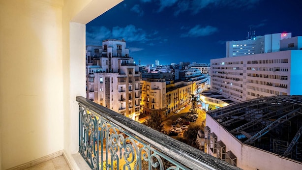 Vista noturna de uma cidade urbana do alto de uma varanda com grade ornamentada, mostrando edifícios altos e um céu limpo.