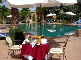 Uma área de refeições à beira da piscina, com uma mesa posta para dois, rodeada por palmeiras e espreguiçadeiras.