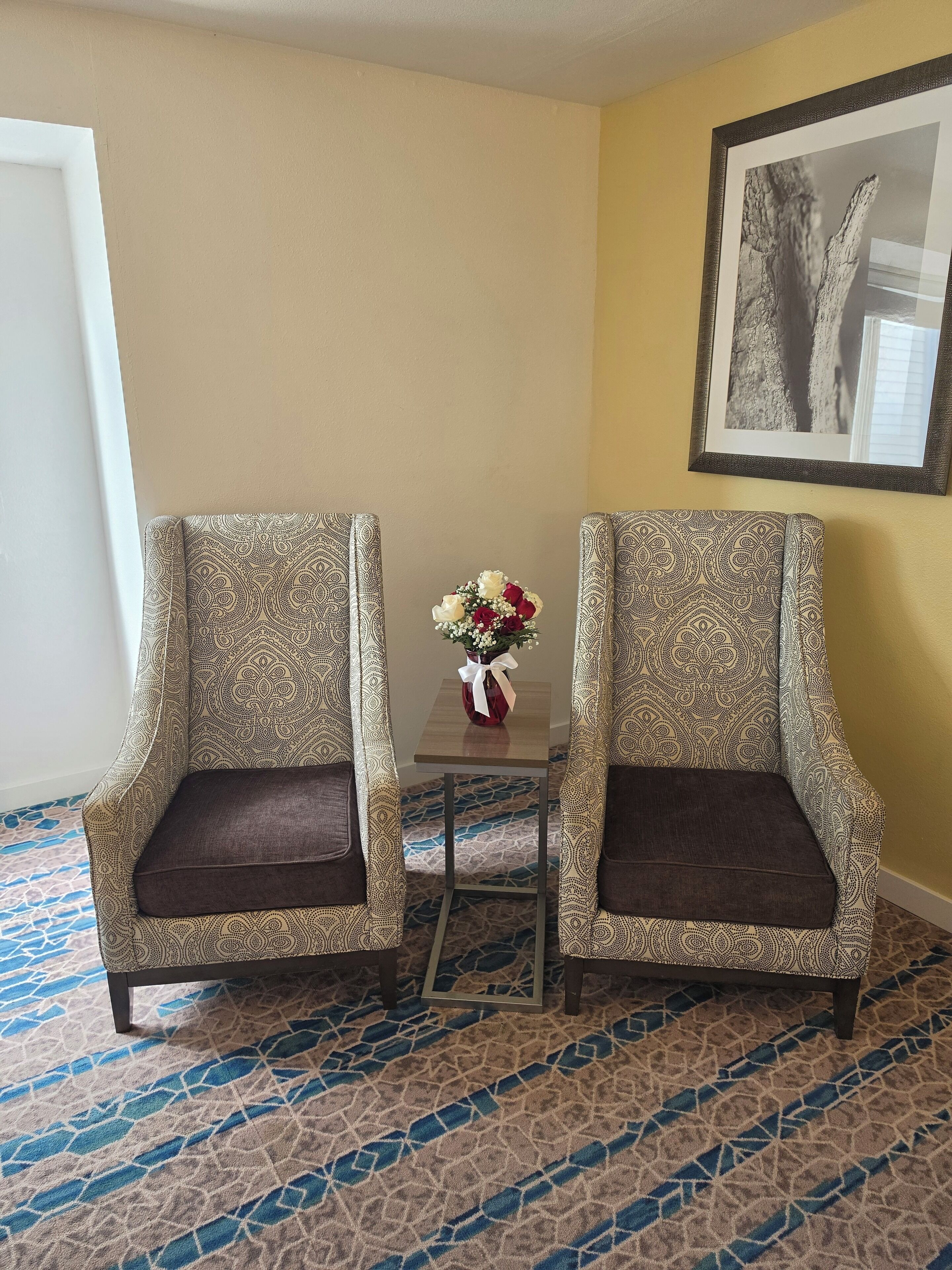 Duas poltronas estampadas com almofadas escuras, uma mesa pequena com um vaso de flores e um quadro na parede.