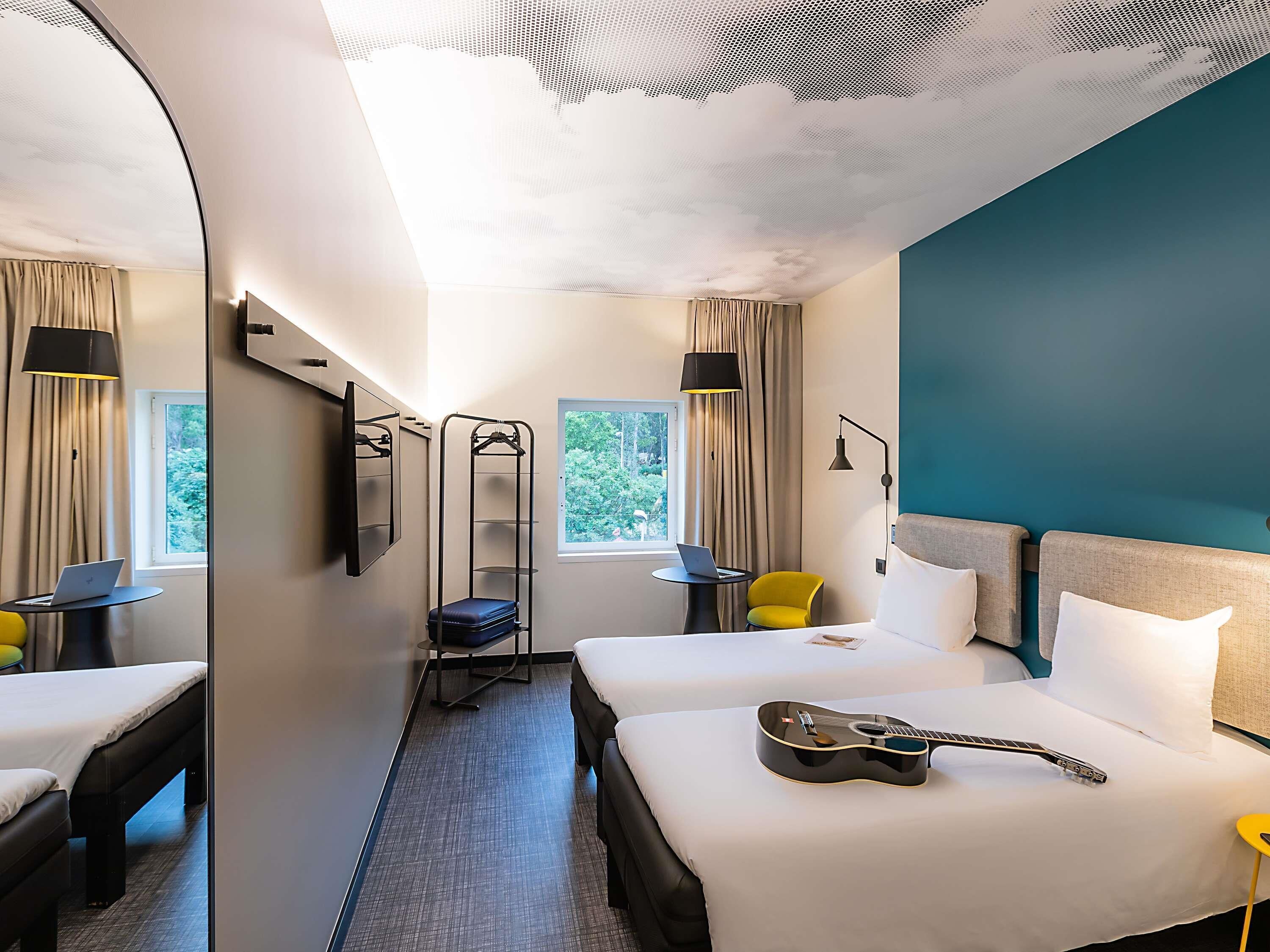Quarto de hotel com duas camas, uma guitarra sobre uma delas, uma escrivaninha com um laptop, uma cadeira, uma estante suspensa e uma janela com cortinas.