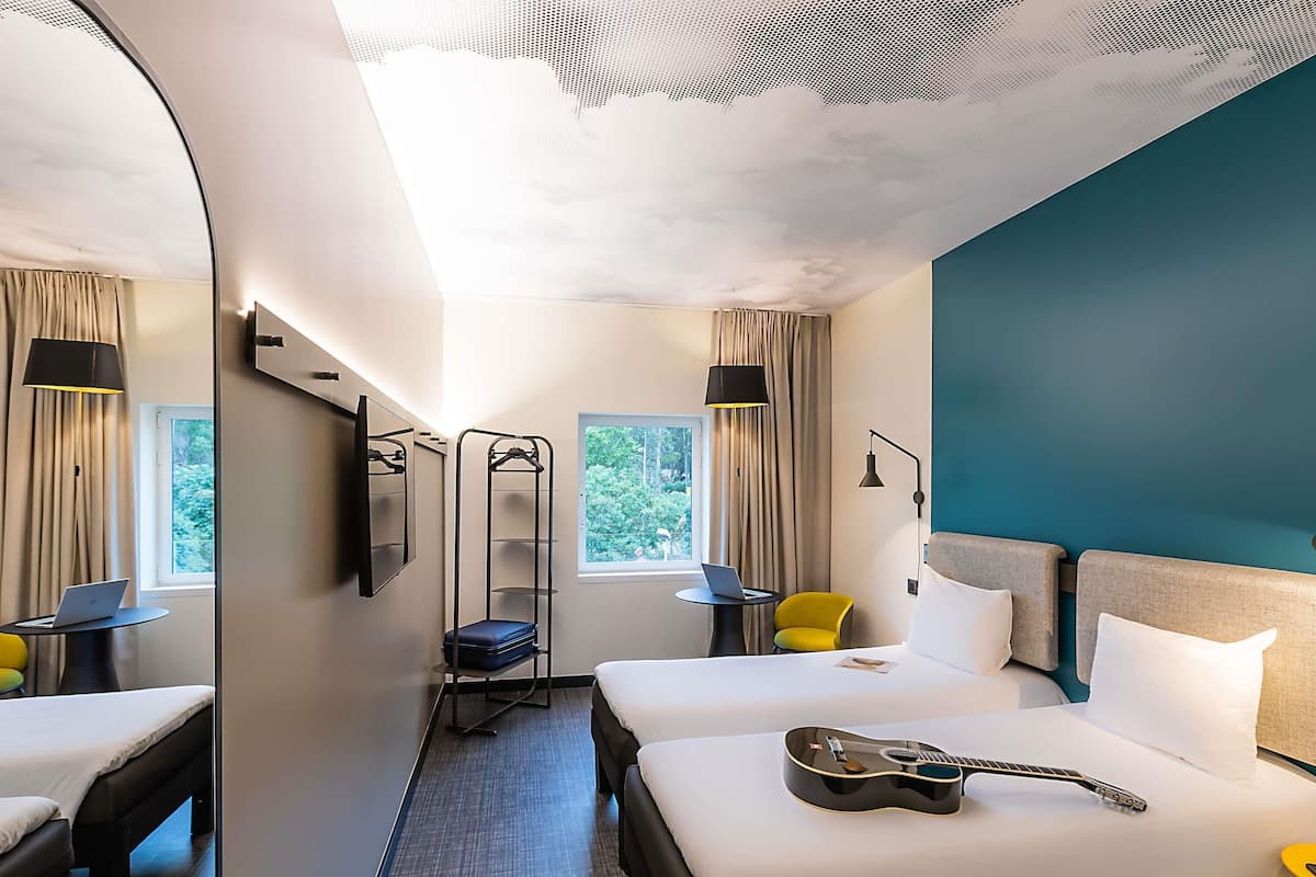Quarto de hotel com duas camas, uma guitarra sobre uma delas, uma escrivaninha com um laptop, uma cadeira, uma estante suspensa e uma janela com cortinas.