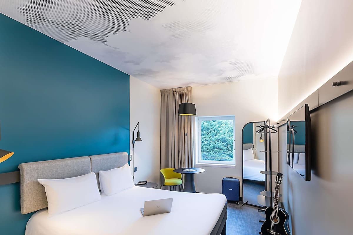Quarto de hotel com cama, escrivaninha, cadeira, guitarra, janela com vista para árvores e teto com padrão geométrico.