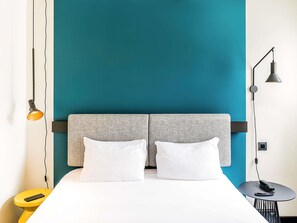 Quarto de hotel moderno com parede azul-esverdeada, cabeceira cinza texturizada, roupa de cama branca, mesa de apoio amarela com um smartphone e uma pequena mesa redonda com um telefone.