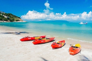 Caiaques alinhados em uma praia, com um barco na água e uma colina ao fundo.