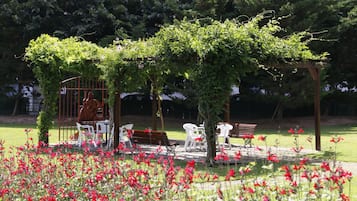 Um jardim com pérgola, bancos e cadeiras sob uma estrutura coberta por trepadeiras.
