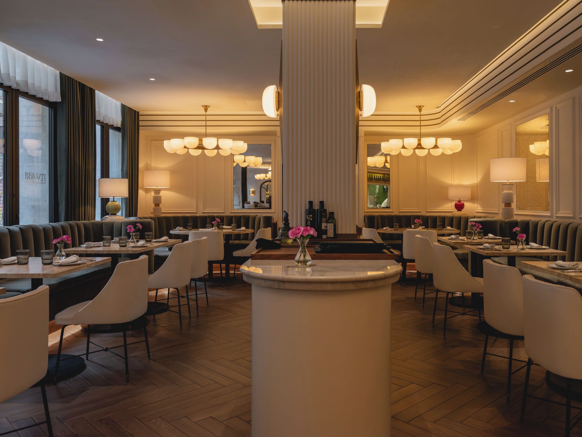 Um restaurante espaçoso com pé-direito alto, lustres modernos e um pilar central. As mesas estão dispostas ao redor do pilar, com assentos em ambos os lados.