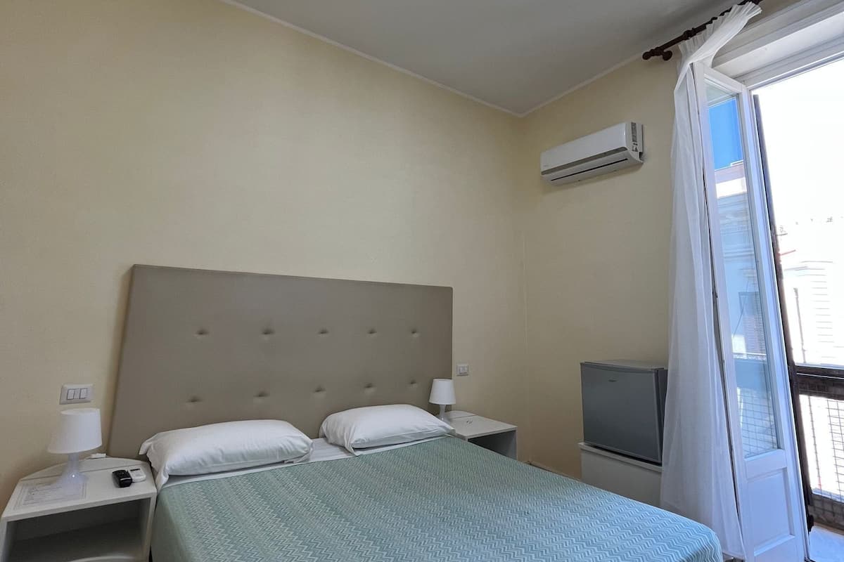 Quarto de hotel com cama, mesas de cabeceira, aparelho de ar condicionado, televisão e janela com cortinas.