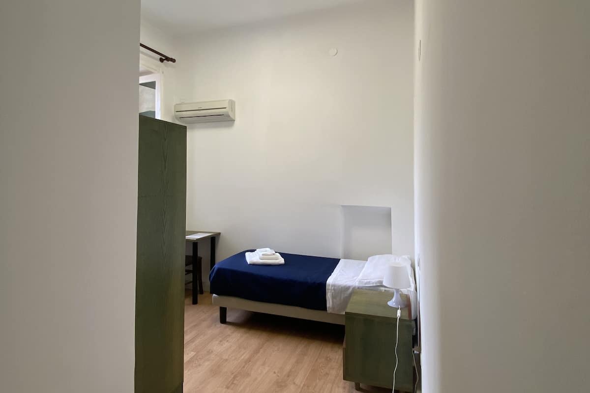 Um quarto pequeno e modesto, com uma cama, uma escrivaninha e um ar condicionado fixado na parede.