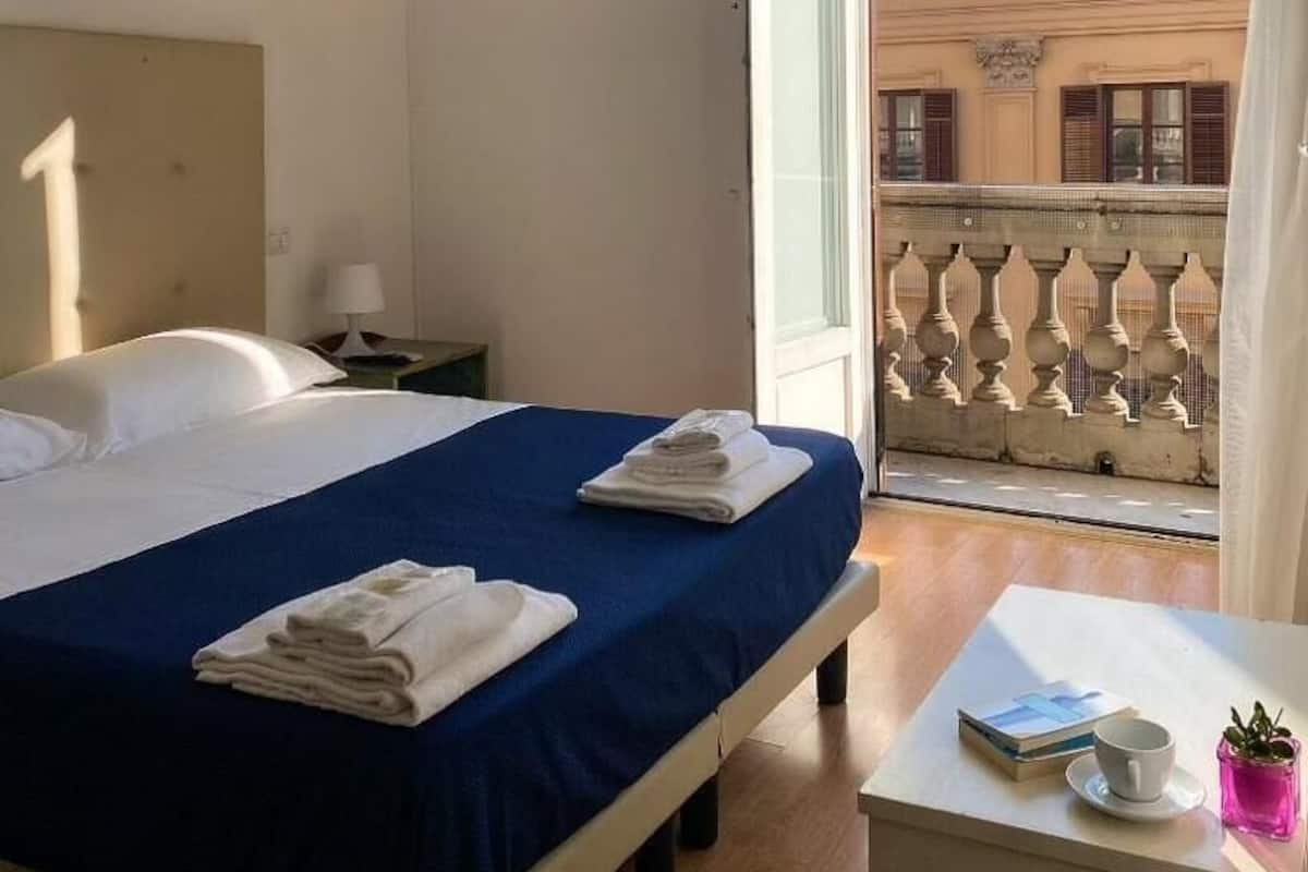 Quarto de hotel com cama, mesa de cabeceira, uma mesa com uma xícara e um livro, e uma varanda com guarda-corpo.